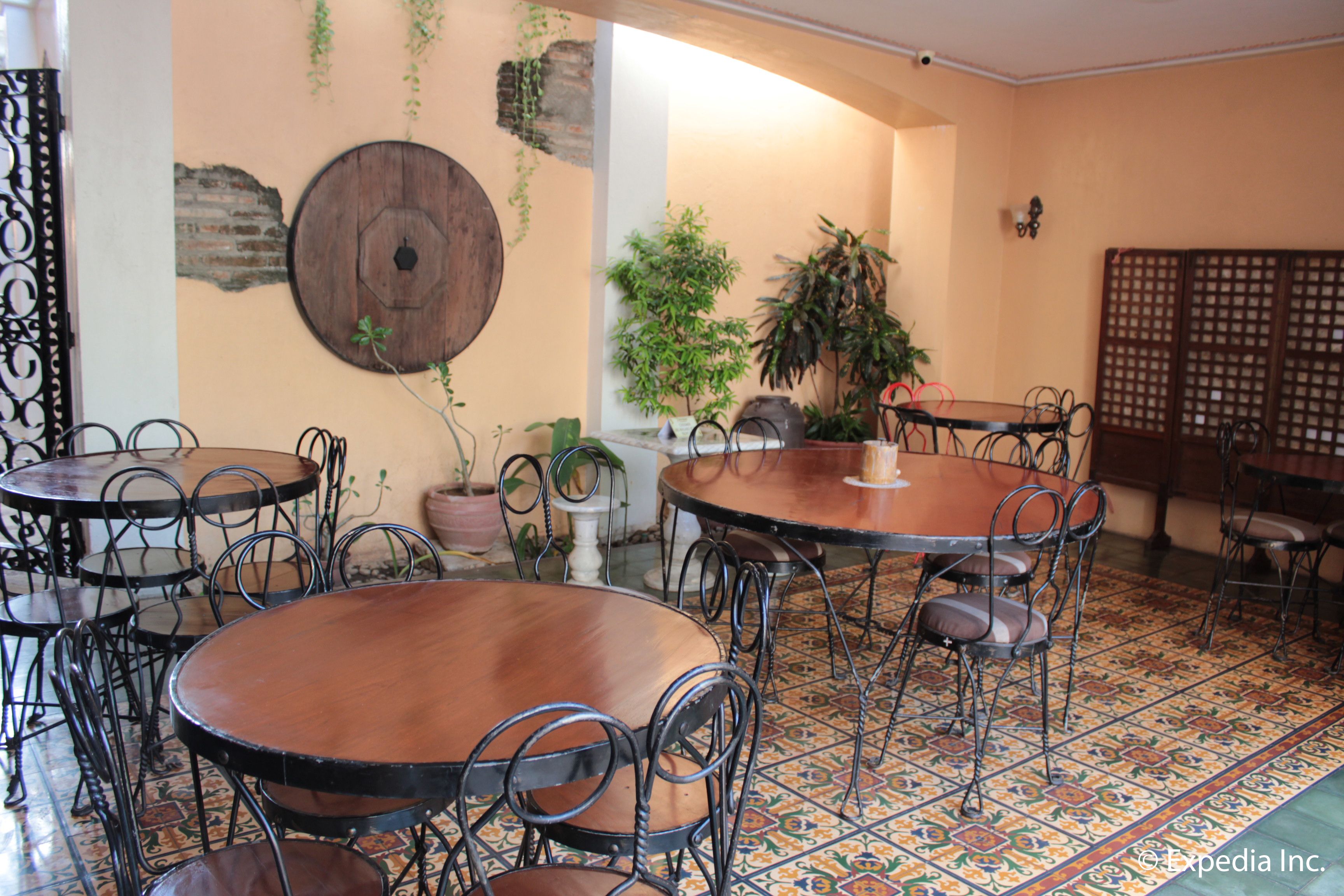 Uma área de refeições com mesas redondas e cadeiras de metal, cercada por plantas em vasos e uma parede decorativa.