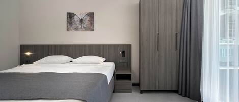 Quarto de hotel moderno com uma cama grande, uma mesinha de cabeceira, um guarda-roupa e uma janela com cortinas.