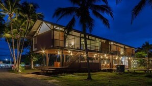 Uma casa moderna de dois andares, com amplas janelas de vidro, um deck de madeira e área de estar externa.