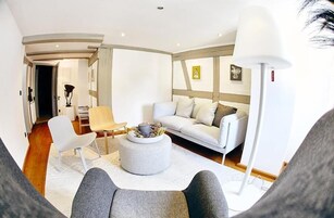 Uma sala de estar moderna com sofá, apoio para os pés e poltronas, uma luminária e um corredor que dá para outro cômodo.