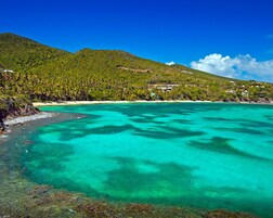 Paisagem costeira com águas turquesa cristalinas, uma praia arenosa e colinas verdes exuberantes ao fundo.