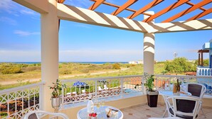 Terraço com móveis de vime branco, uma mesa com garrafa, copos e flor, e vista para o mar e um moinho de vento.