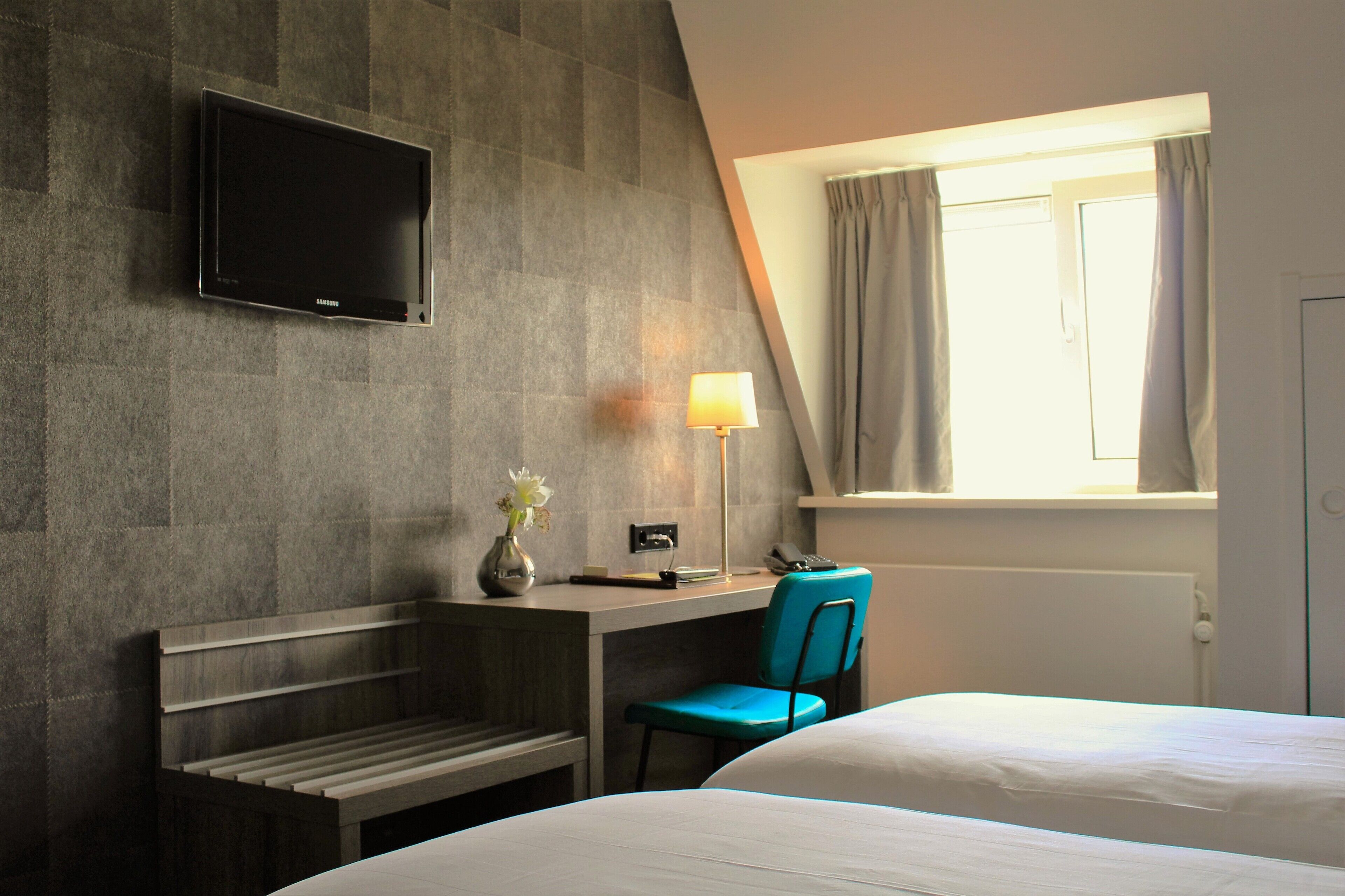 Quarto de hotel com uma cama, uma escrivaninha com cadeira, televisão fixada na parede, janela com cortinas e um vaso de flores na escrivaninha.