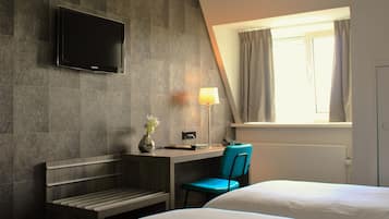 Quarto de hotel com uma cama, uma escrivaninha com cadeira, televisão fixada na parede, janela com cortinas e um vaso de flores na escrivaninha.