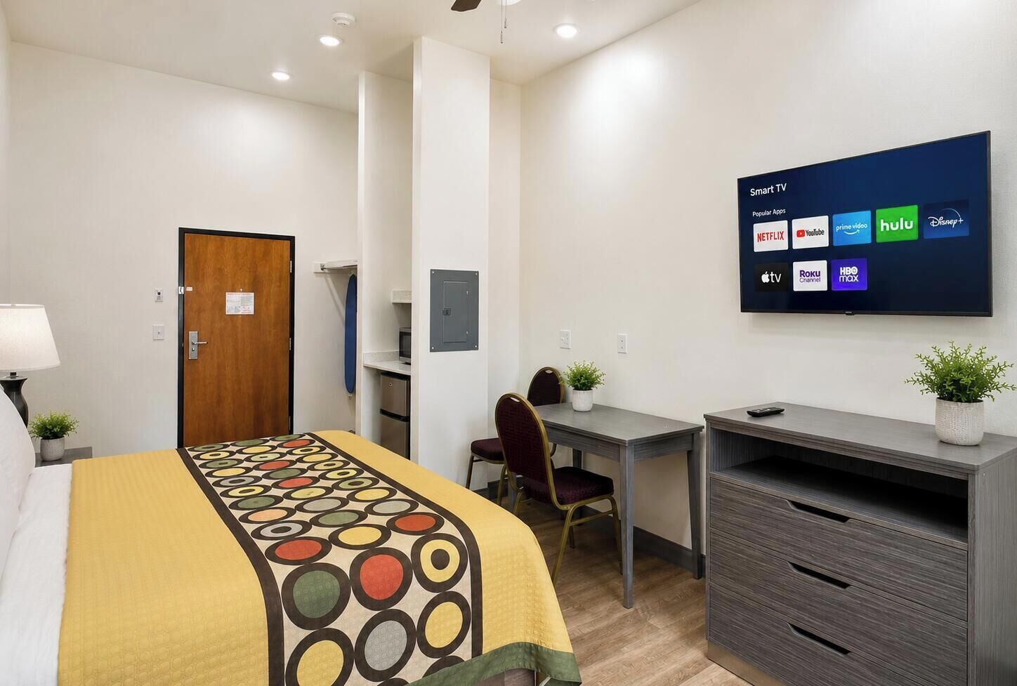 Quarto de hotel com cama, escrivaninha, cadeira, TV exibindo serviços de streaming e uma pequena planta.