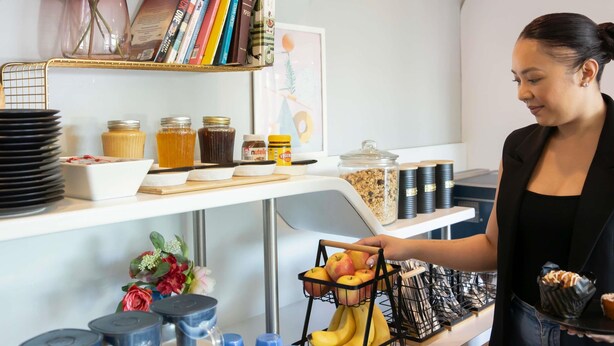 Uma cozinha com alguém servindo comida, uma estante de livros e diversos utensílios na bancada.