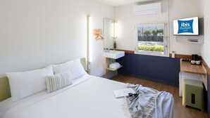 Quarto de hotel com cama, mesa de cabeceira com luminária, janela com vista, pia e uma televisão exibindo "ibis budget hotels".