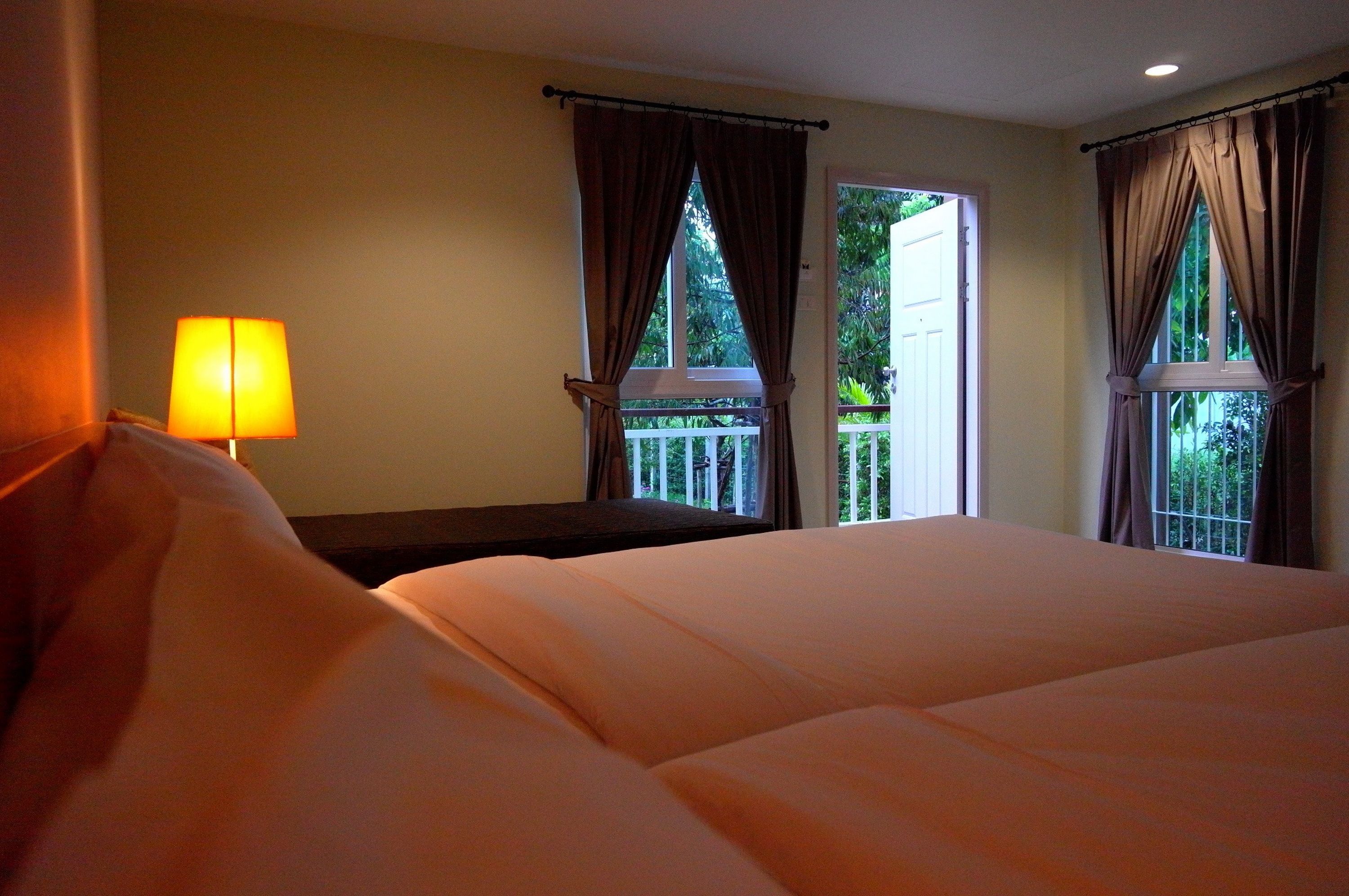 Quarto de hotel com uma cama grande, uma mesa de cabeceira, uma luminária e uma varanda com cortinas.