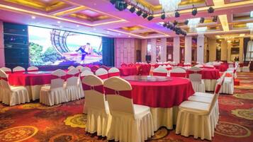 Salão de banquetes com mesas redondas, cadeiras brancas e palco com uma tela grande.