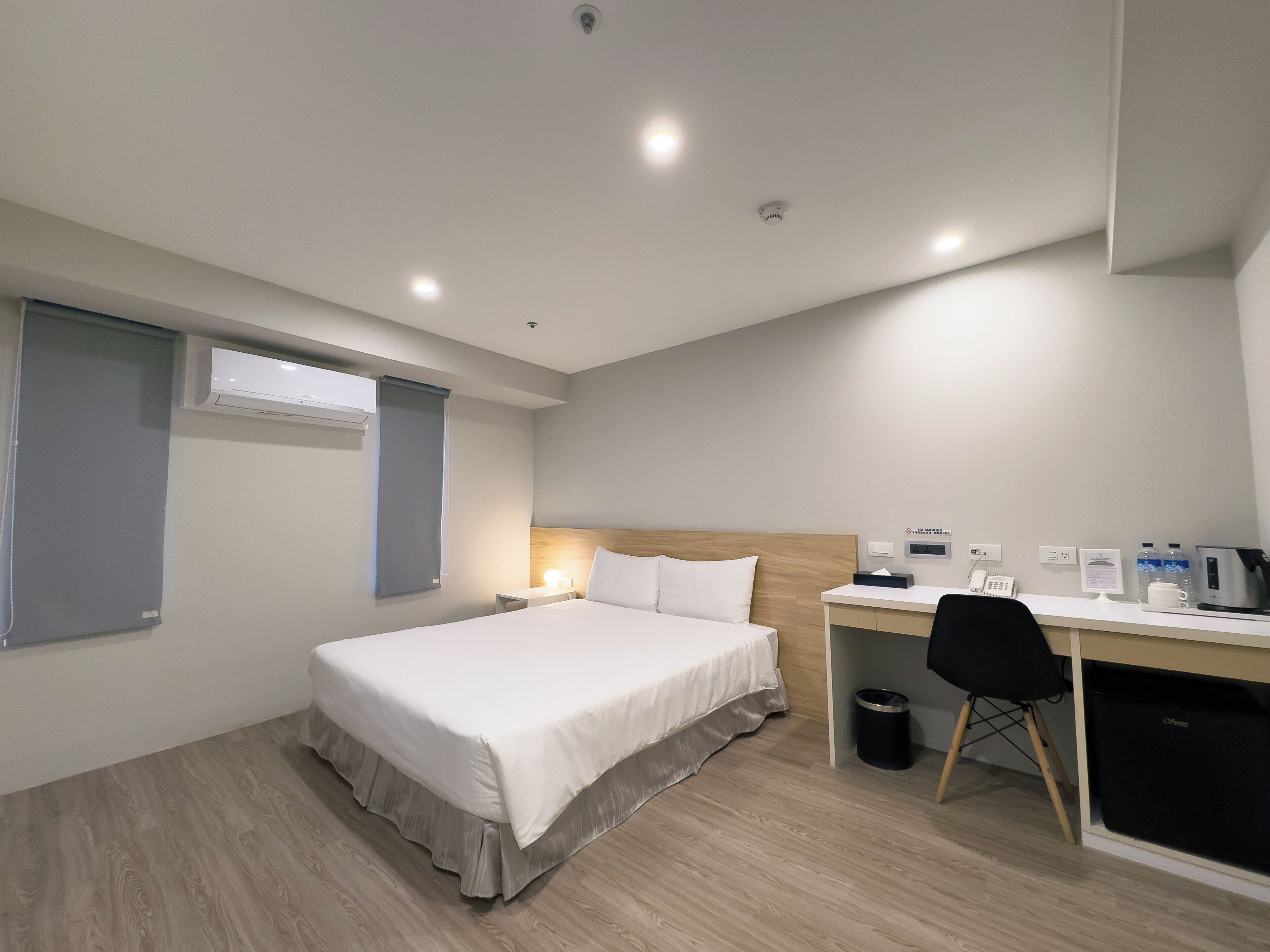 Quarto de hotel com cama, escrivaninha, cadeira e uma pequena lixeira.