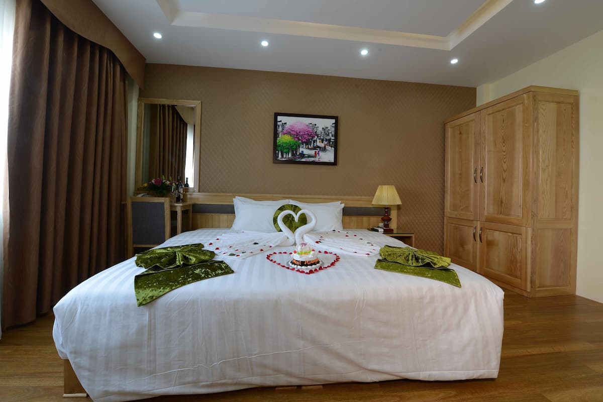 Cama bem arrumada com decoração em formato de coração e almofadas em forma de cisne, guarda-roupa de madeira e quadro na parede.