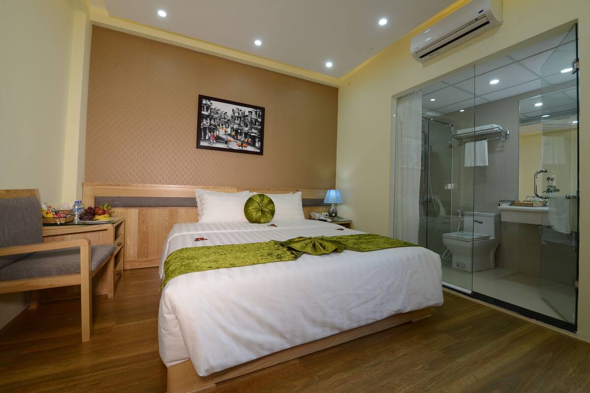 Quarto de hotel com cama, secretária, cadeira, mesa de cabeceira, luminária, quadro na parede e banheiro com box de vidro.