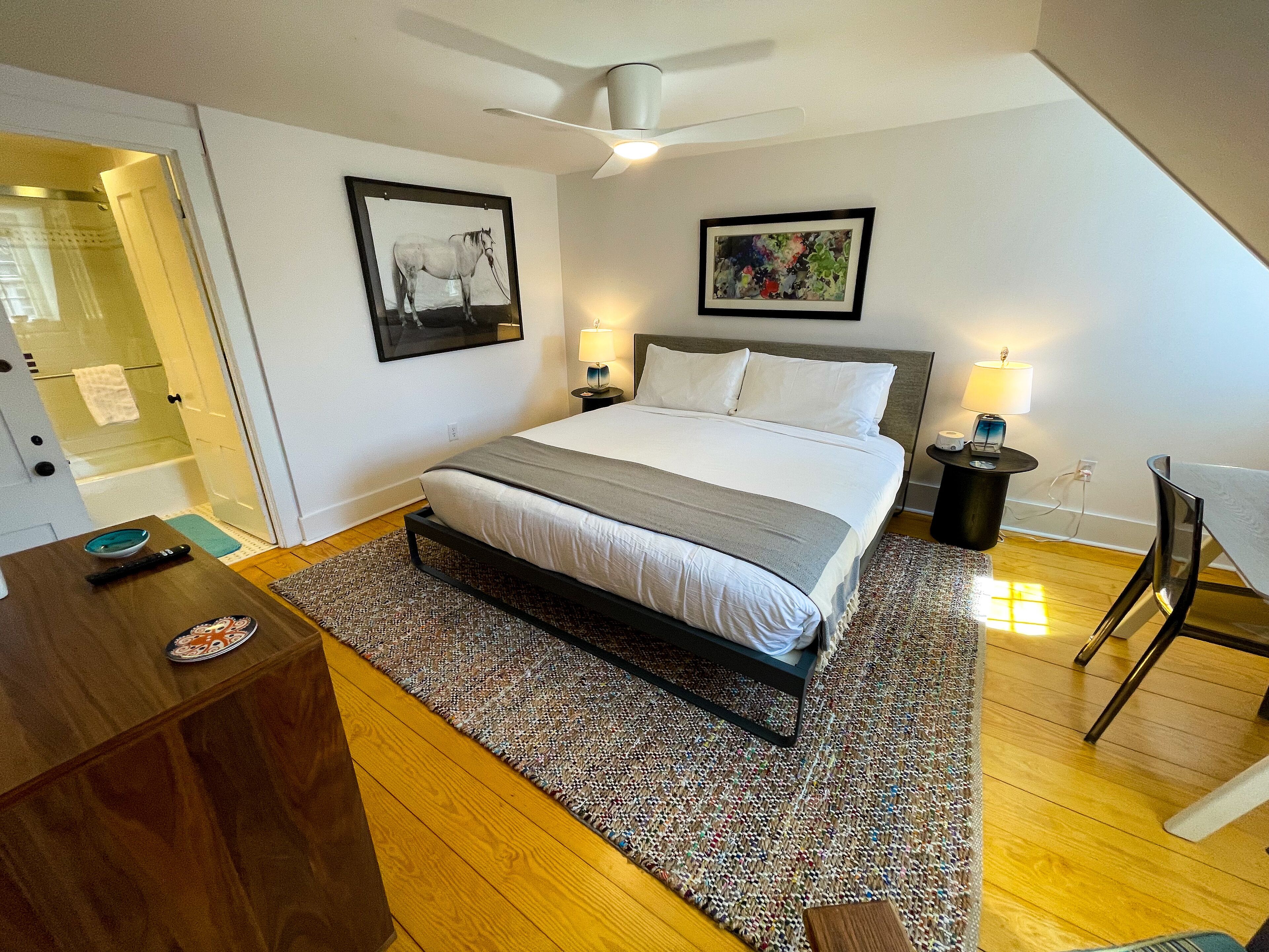 Quarto com cama, mesas de cabeceira, escrivaninha, cadeira, ventoinha no teto e duas imagens emolduradas na parede.