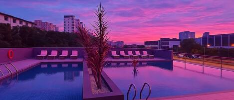 Piscina no terraço com espreguiçadeiras e vista para o horizonte urbano ao entardecer.