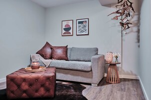 Uma sala de estar moderna com um sofá cinzento, almofadas castanhas e cinzentas, um banquinho castanho e uma pequena mesa lateral de madeira com um vaso e flores.