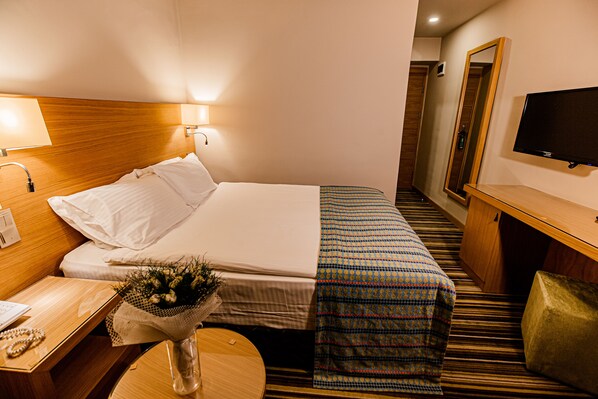 Quarto de hotel com cama bem arrumada, mesa de cabeceira com abajur, pequena mesa com um vaso de flores, espelho e televisão sobre um suporte.