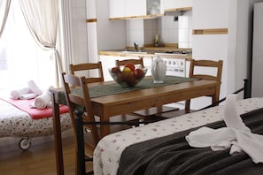Quarto de hotel com uma mesa de jantar de madeira, uma tigela de frutas, um jarro branco e uma cama com colcha florida.