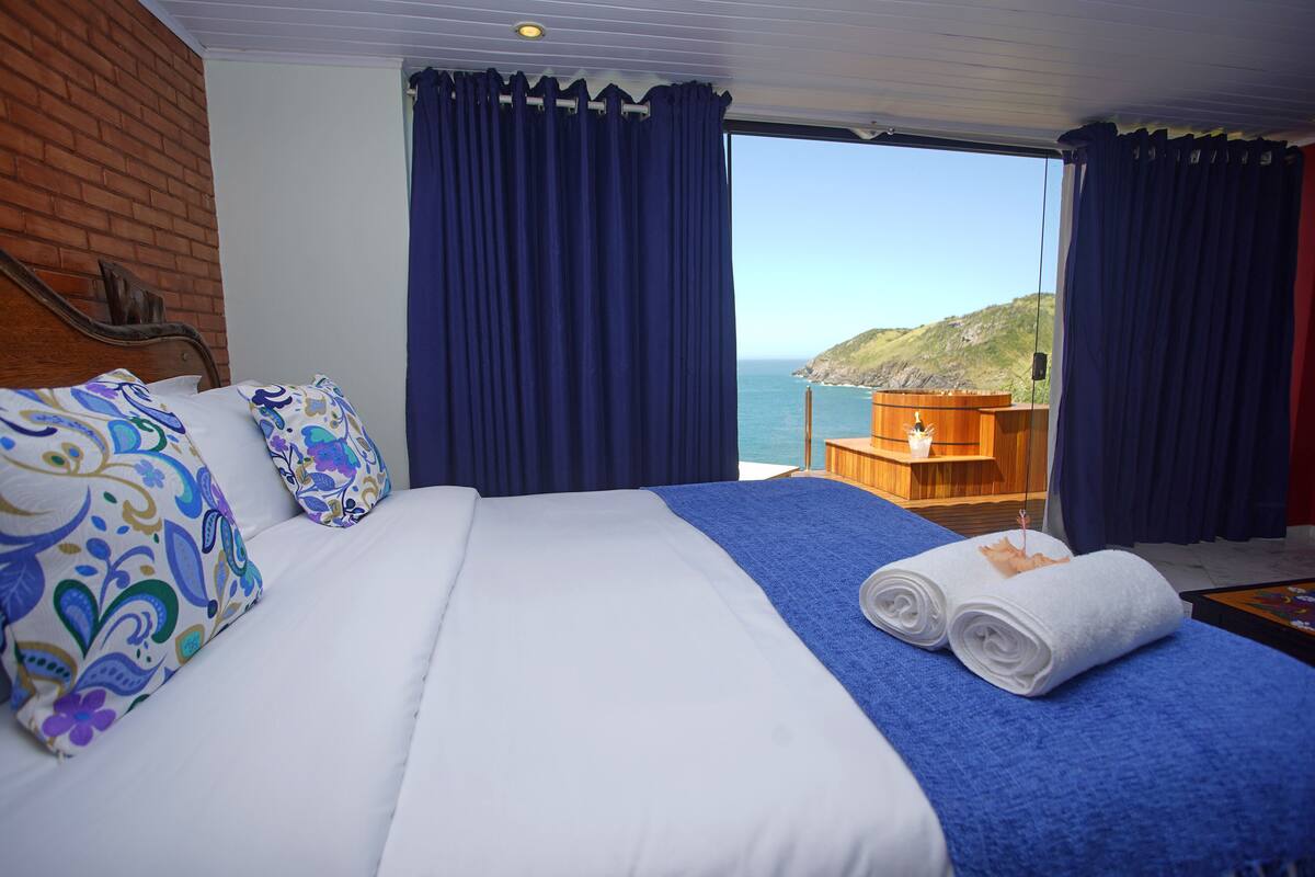 Quarto de hotel com uma cama grande, cortinas azuis e vista para o mar.