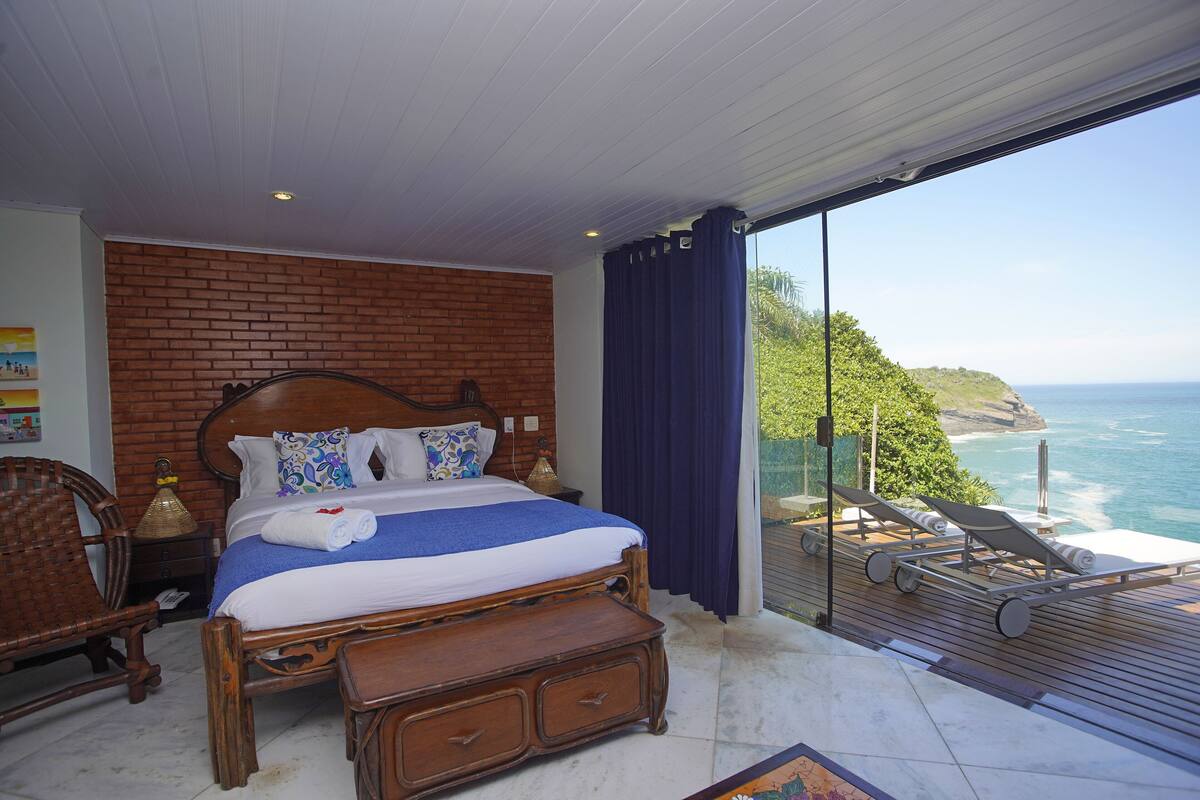 Quarto com cama grande, cabeceira de madeira, mesa de cabeceira, cadeira de vime e vista para o oceano.