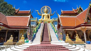 Um templo com uma grande estátua dourada no topo de uma escadaria, rodeado por estátuas menores e decorações detalhadas.
