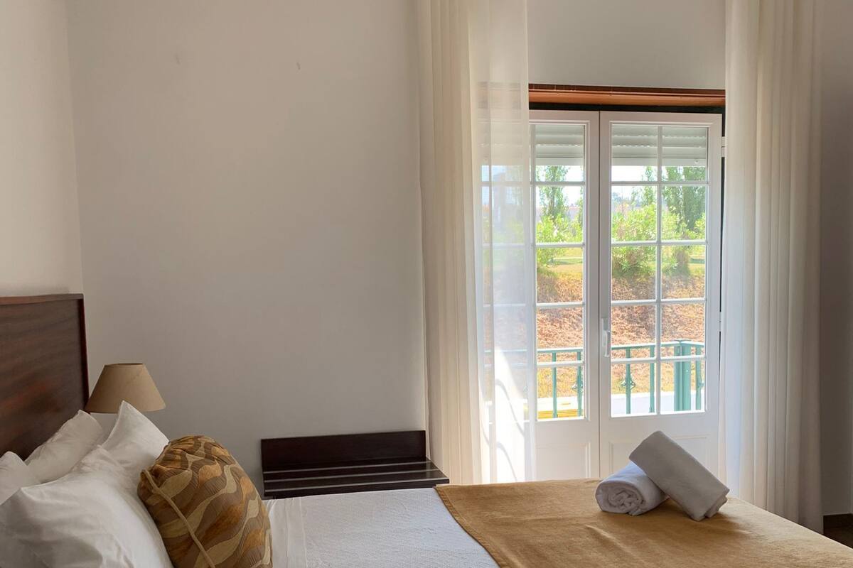 Quarto de hotel com cama, cabeceira de madeira, janela com cortinas e vista para o exterior.