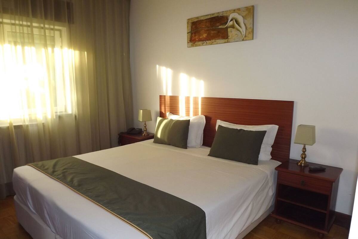Quarto de hotel com uma cama grande, duas mesas de cabeceira com luminárias, um criado-mudo e um quadro na parede.