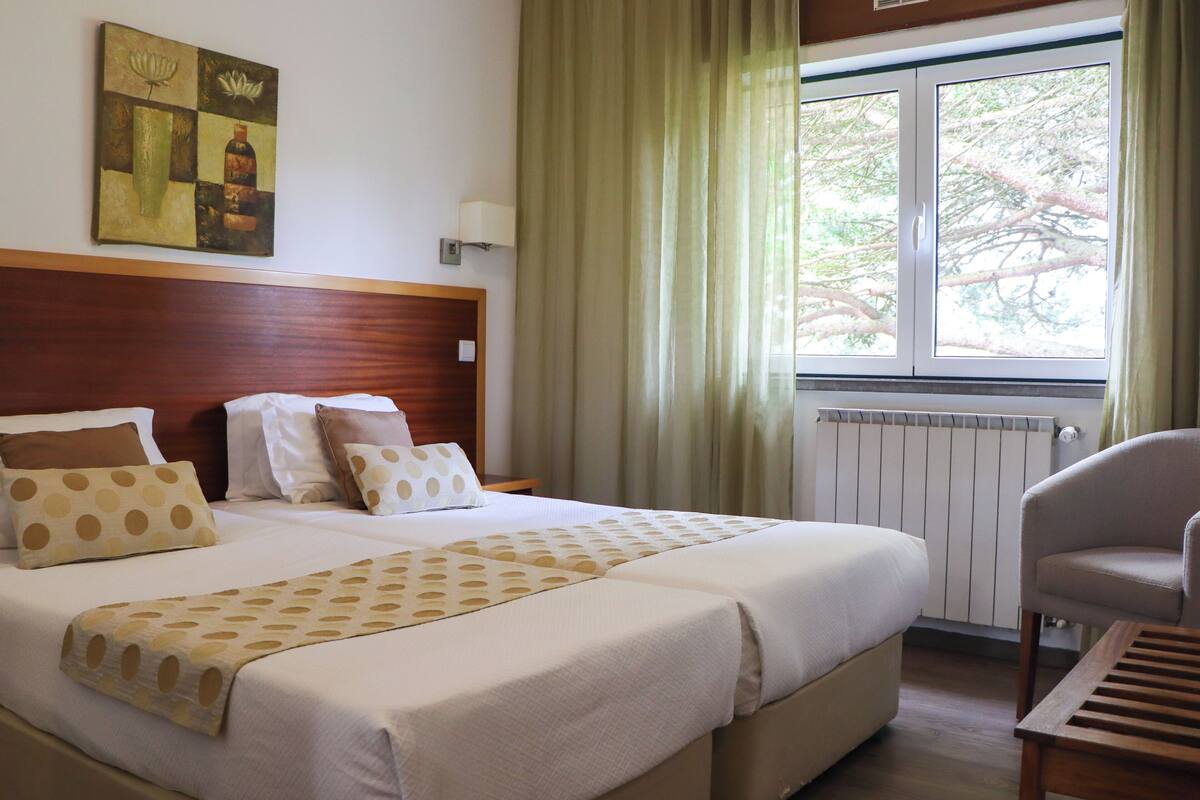 Quarto de hotel com uma cama grande, uma cadeira, janela com cortinas e luminária na parede.