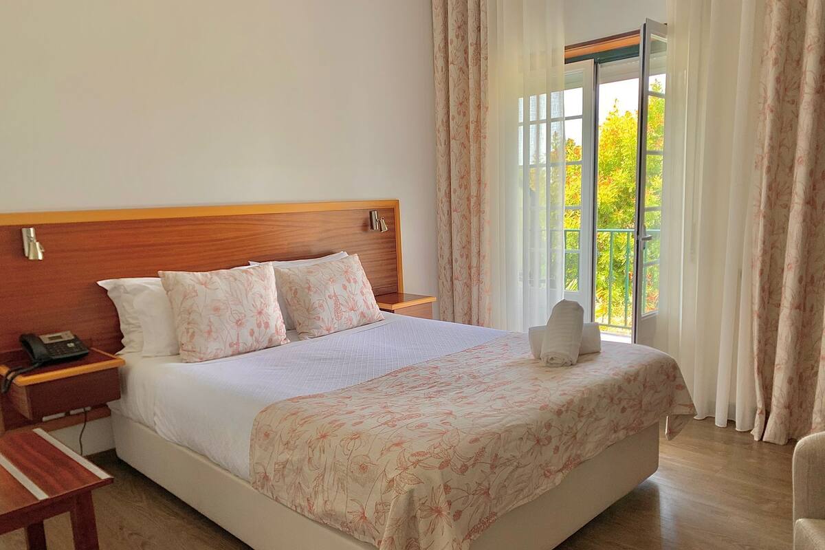 Quarto de hotel com cabeceira de madeira, cama grande com roupa de cama floral, mesa de cabeceira com telefone e vista para o verde pela janela.