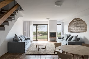 Uma sala de estar moderna com um conjunto de sofás, uma mesa de centro redonda, uma mesa de jantar com cadeiras, uma lareira e vista para o exterior.