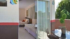 Quarto de hotel com cama, uma mesinha e uma cadeira. Há uma porta de vidro deslizante que dá para uma varanda com uma planta em vaso.