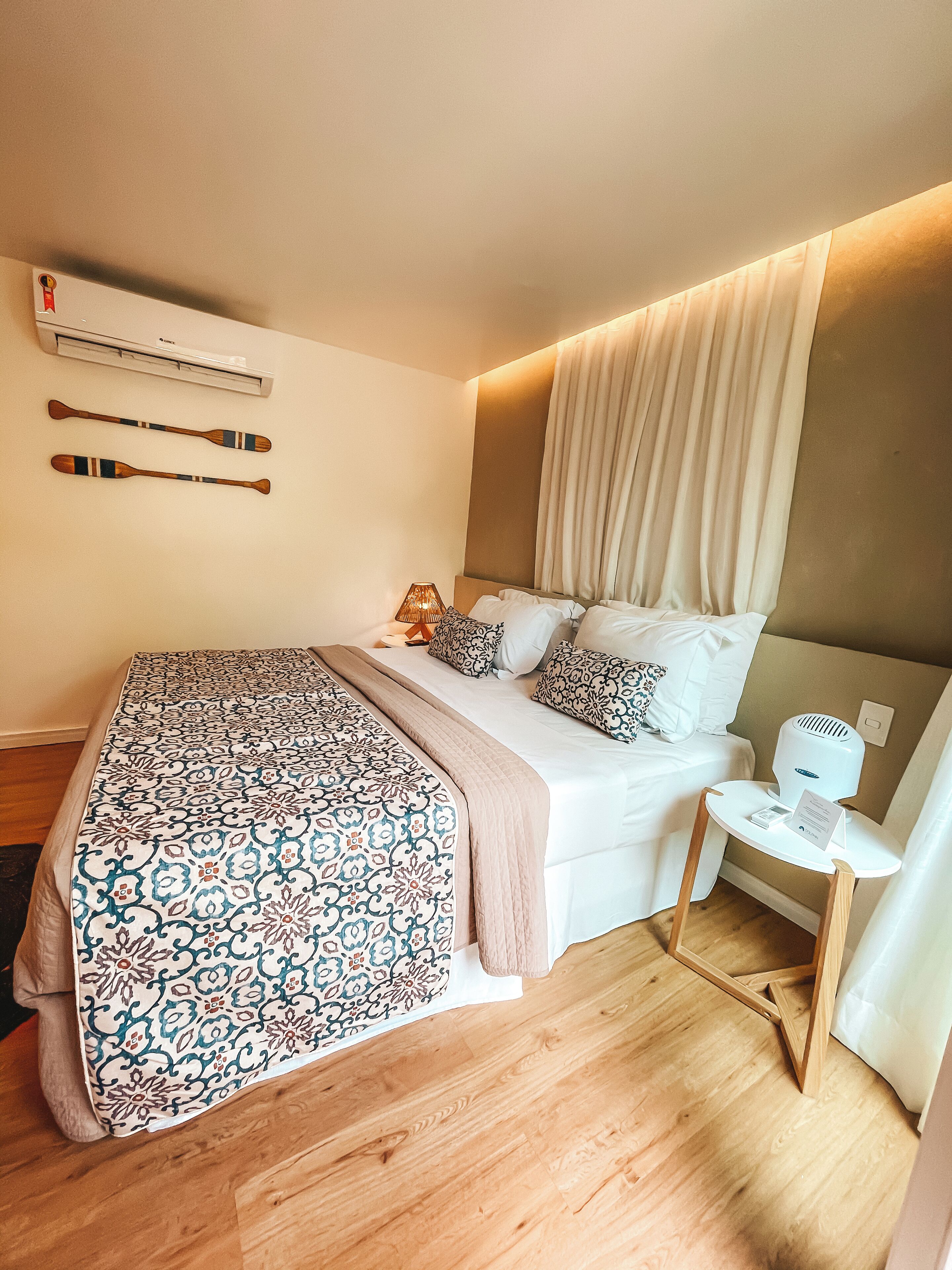 Quarto de hotel moderno com uma cama grande, uma mesa de cabeceira com luminária e ar-condicionado instalado no teto.