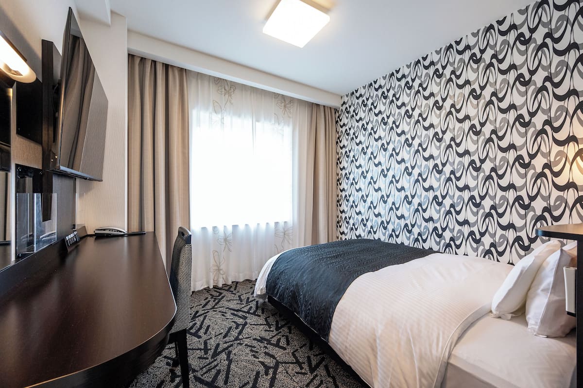 Quarto de hotel com cama, escrivaninha, cadeira, televisão e parede estampada.
