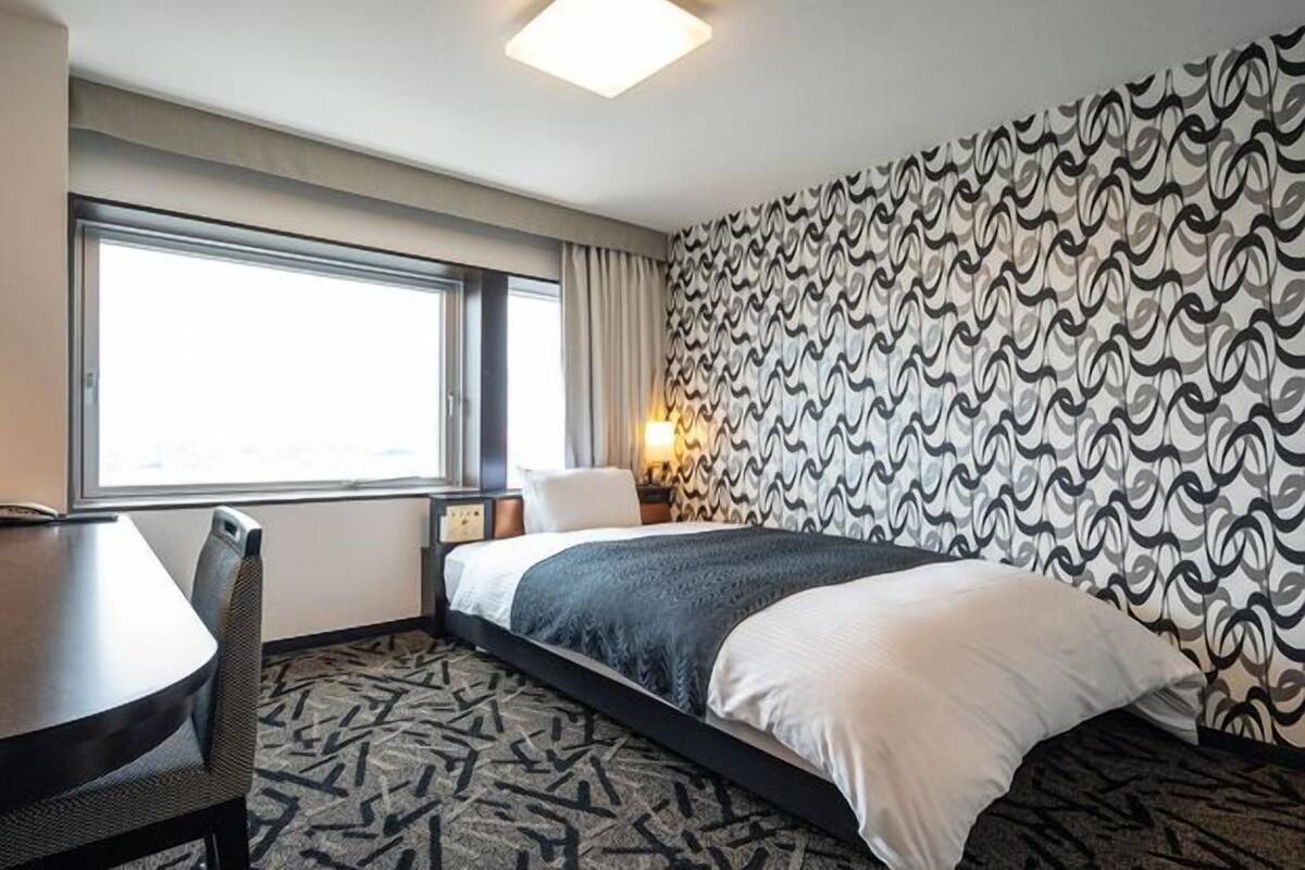 Quarto de hotel com cama, escrivaninha, cadeira, janela e parede com padrão.