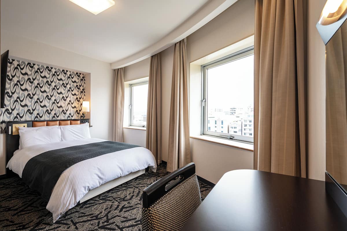 Quarto de hotel com cama, escrivaninha, cadeira, janela com cortinas e parede estampada.