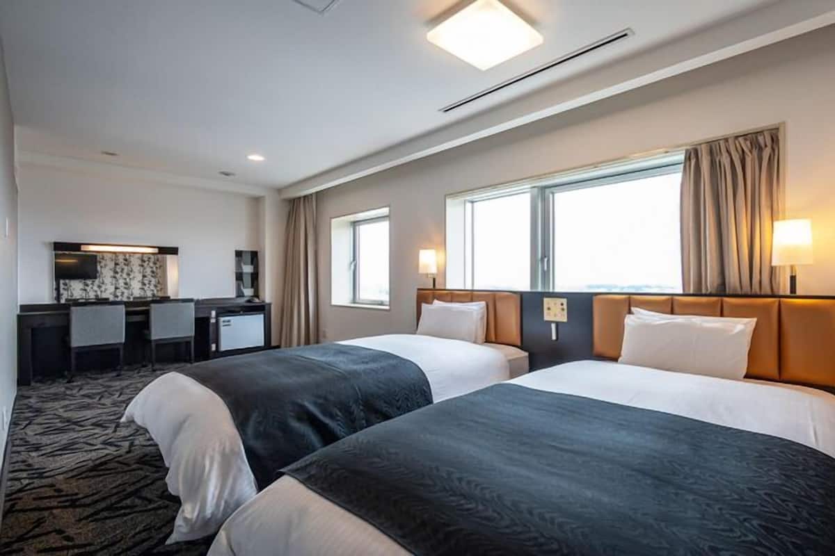 Quarto de hotel com duas camas, uma escrivaninha e uma janela grande.