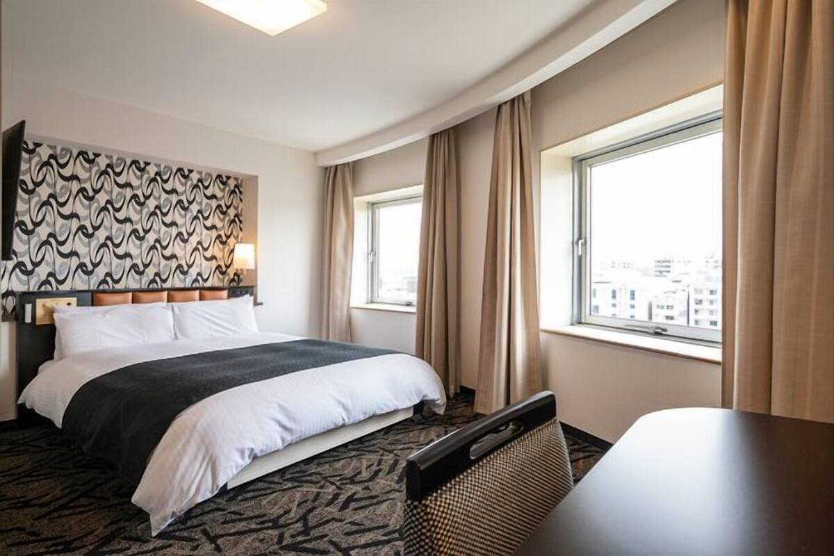 Quarto de hotel com cama, escrivaninha, janela com cortinas e vista para a cidade.