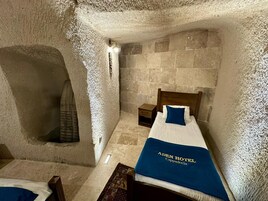 Quarto com parede de pedra, cama com colcha azul, mesa de cabeceira de madeira e cômoda pequena.
