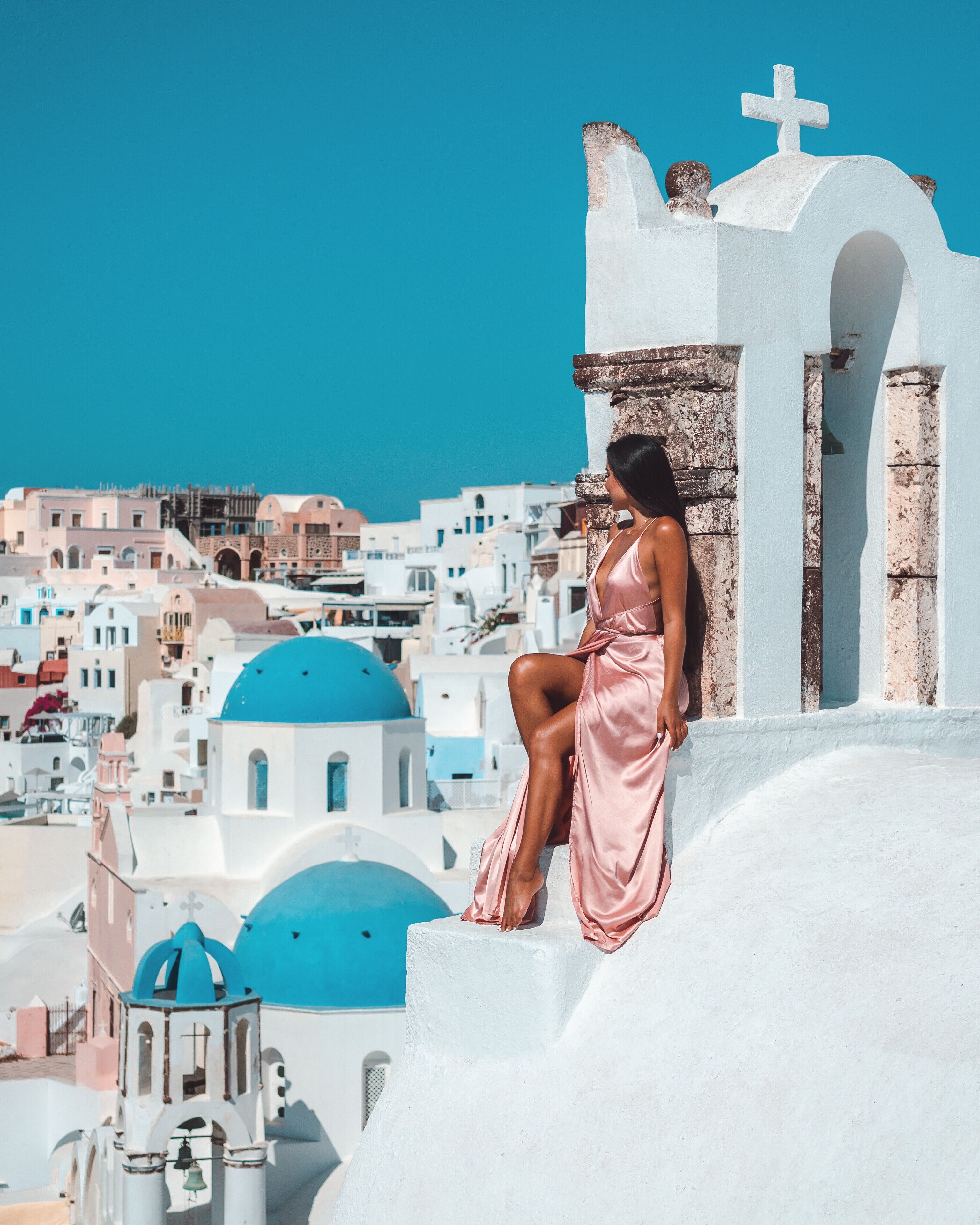 Mulher com vestido rosa sentada em estrutura branca com cruz no topo, com vista para uma cidade com edifícios brancos e cúpulas azuis.