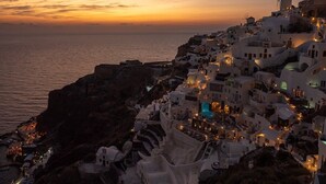 Uma vila costeira com edifícios brancos, um moinho de vento e um pôr do sol sobre o mar.