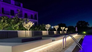 Uma área de piscina com um caminho de pedra, espaços para sentar e um edifício moderno ao fundo.