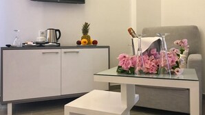 Uma sala de estar moderna com uma TV de tela plana na parede, um armário branco com cafeteira e frutas, uma mesa de vidro com flores cor-de-rosa e um sofá cinza.