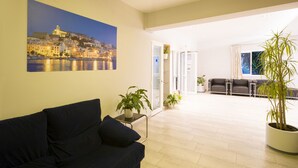 Uma sala de estar com um sofá azul escuro, uma grande pintura de uma paisagem costeira e um vaso com planta.