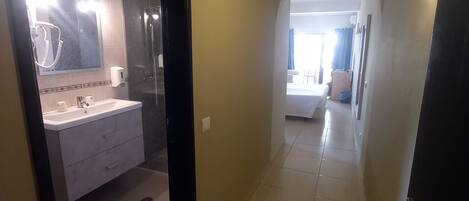 Um corredor de hotel com banheiro equipado com pia, espelho e chuveiro. O corredor dá acesso a um quarto com cama e janela.