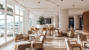 Um lobby moderno de hotel com uma área de estar ampla, grandes janelas com vista para o cais e um arranjo de plantas no centro.
