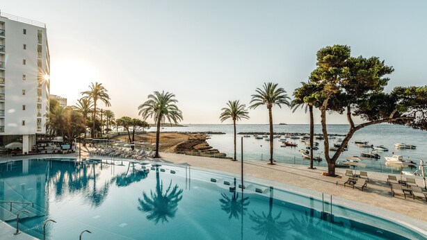 Piscina com espreguiçadeiras e palmeiras, com vista para uma costa rochosa e barcos.