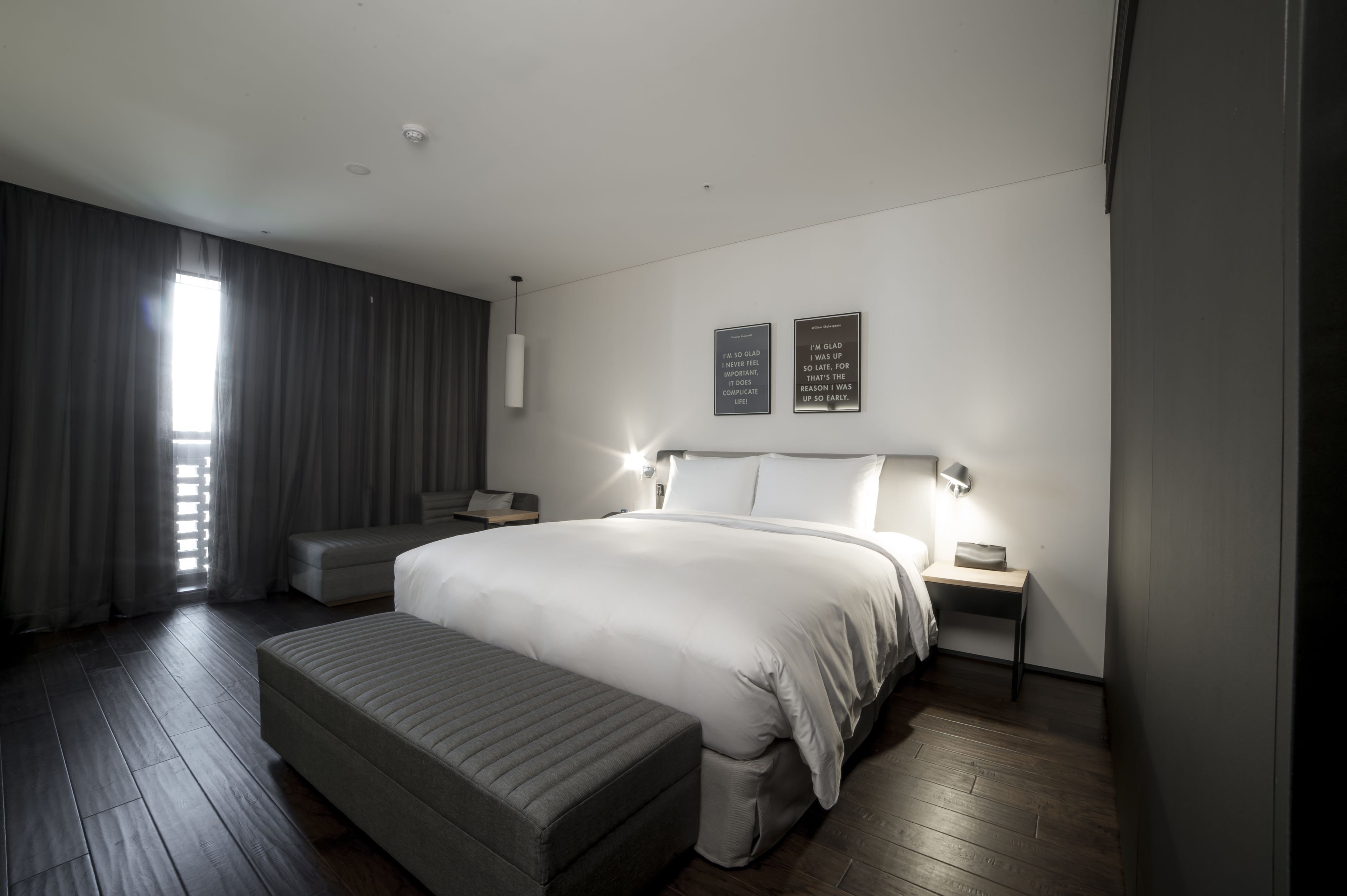 Quarto moderno com uma cama grande, uma mesa de cabeceira, um banco e duas luminárias fixadas na parede.