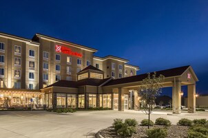 Um hotel com vários andares, um logotipo vermelho chamativo, entrada coberta e um acesso por um caminho arborizado.