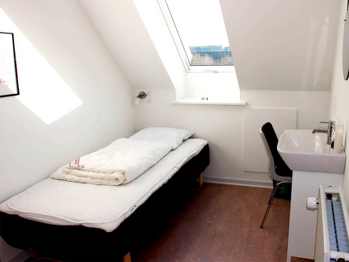 Quarto pequeno com uma cama de solteiro, claraboia, pia branca e uma cadeira.