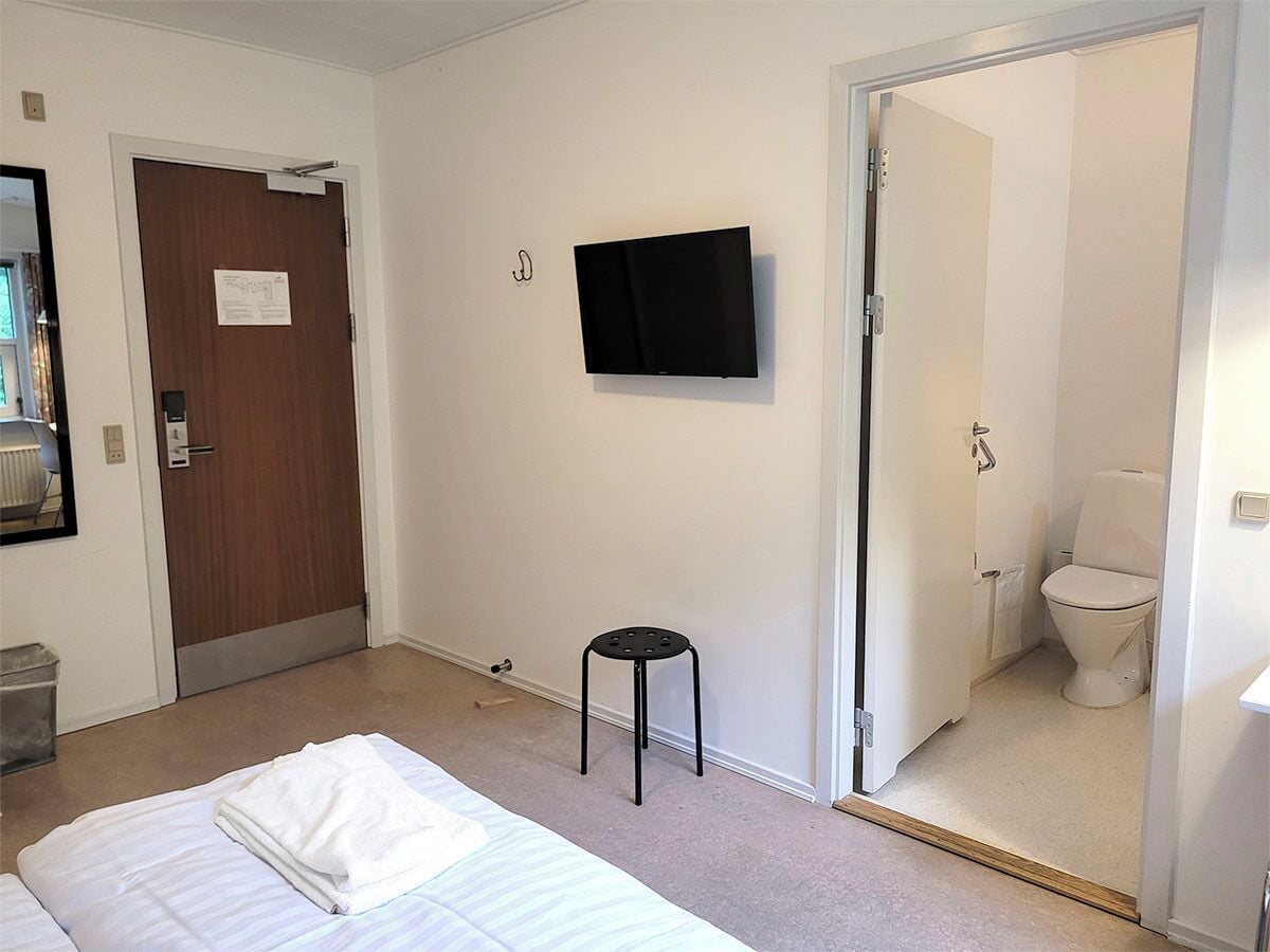 Quarto de hotel com uma cama, um banquinho pequeno, uma televisão fixada na parede e uma porta para o banheiro.