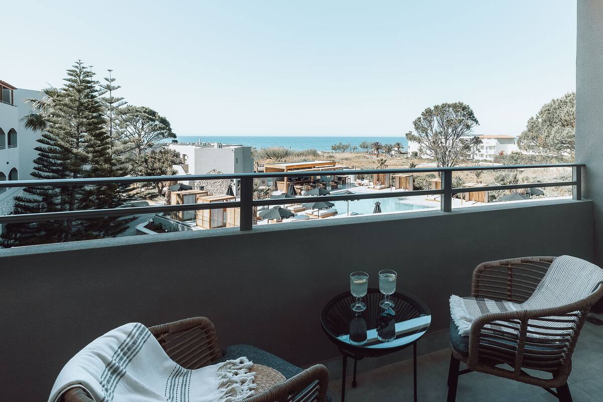 Uma varanda com cadeiras de vime, uma pequena mesa com copos e vista para o mar e edifícios.